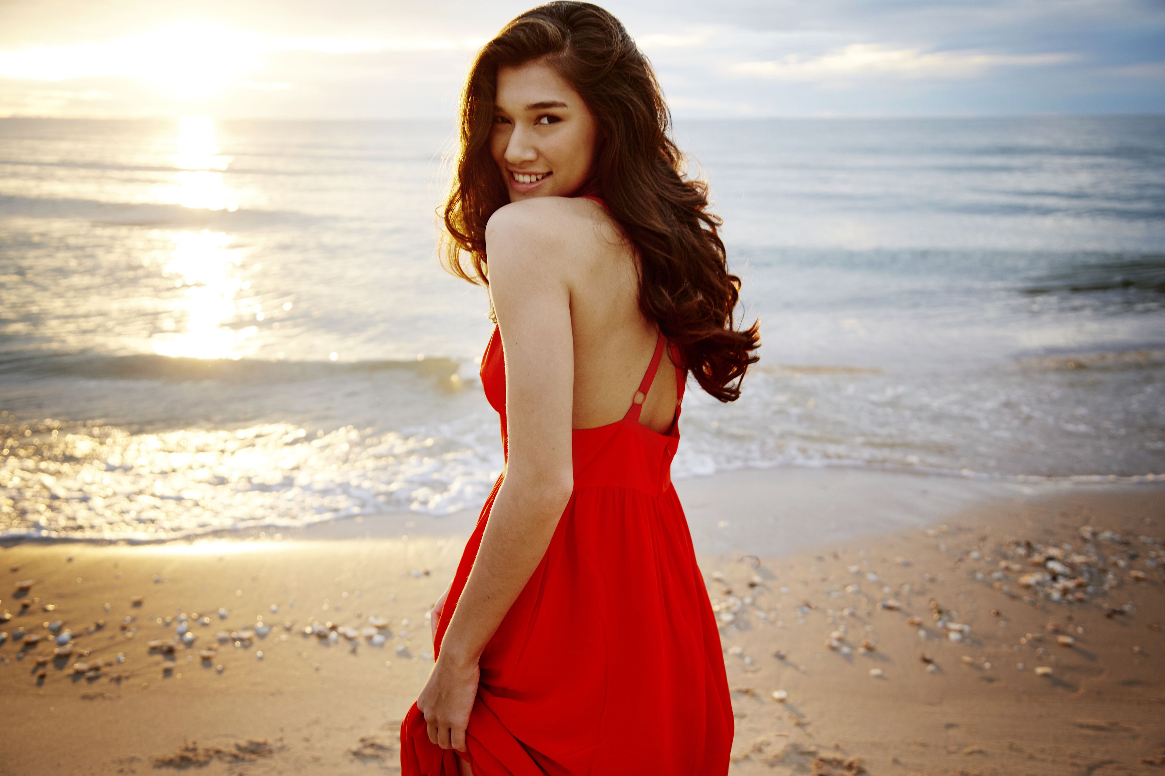 Uma mulher de vestido vermelho em pé numa praia, com o oceano ao fundo.