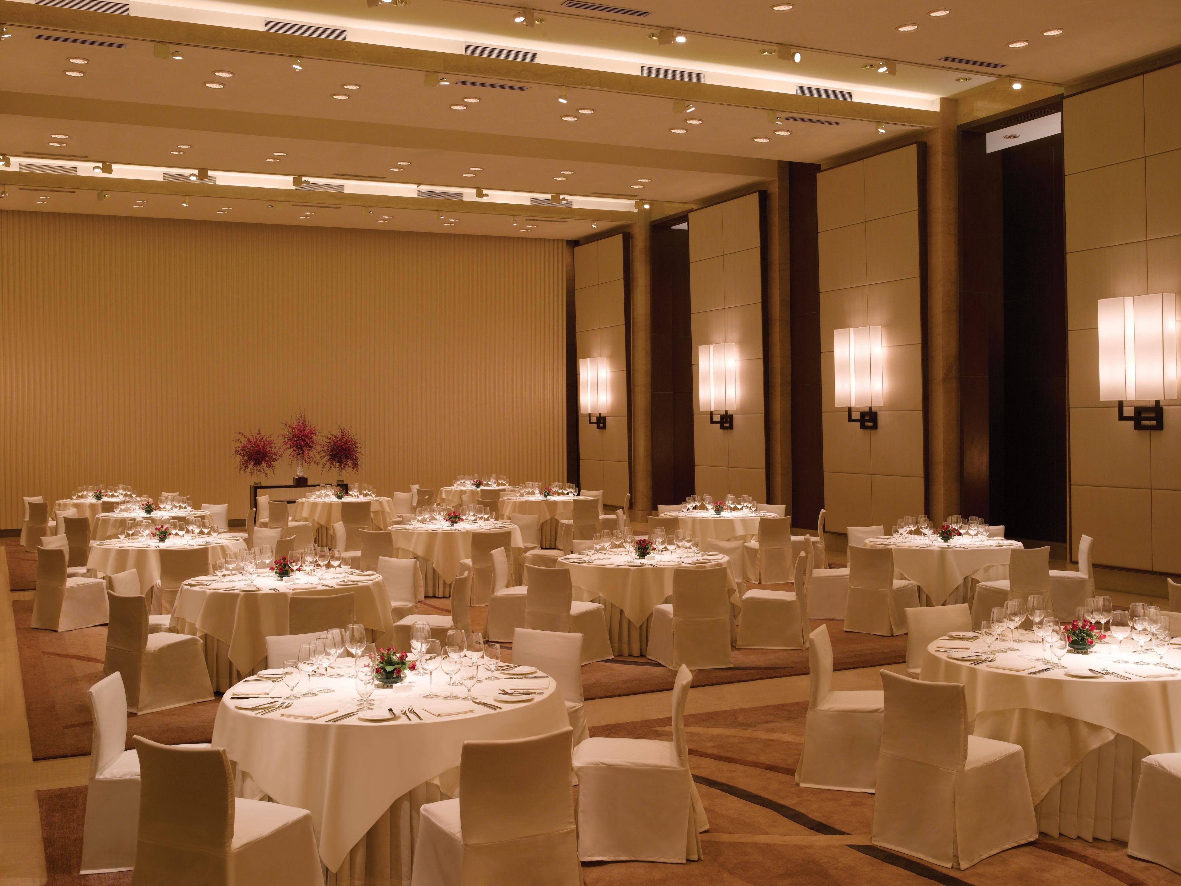 Salão de banquetes com mesas redondas preparadas para refeições, cadeiras brancas e luminárias fixadas na parede.