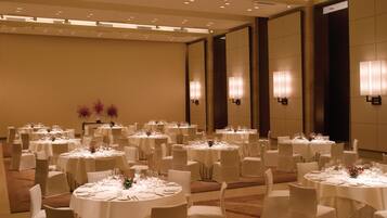 Salão de banquetes com mesas redondas preparadas para refeições, cadeiras brancas e luminárias fixadas na parede.