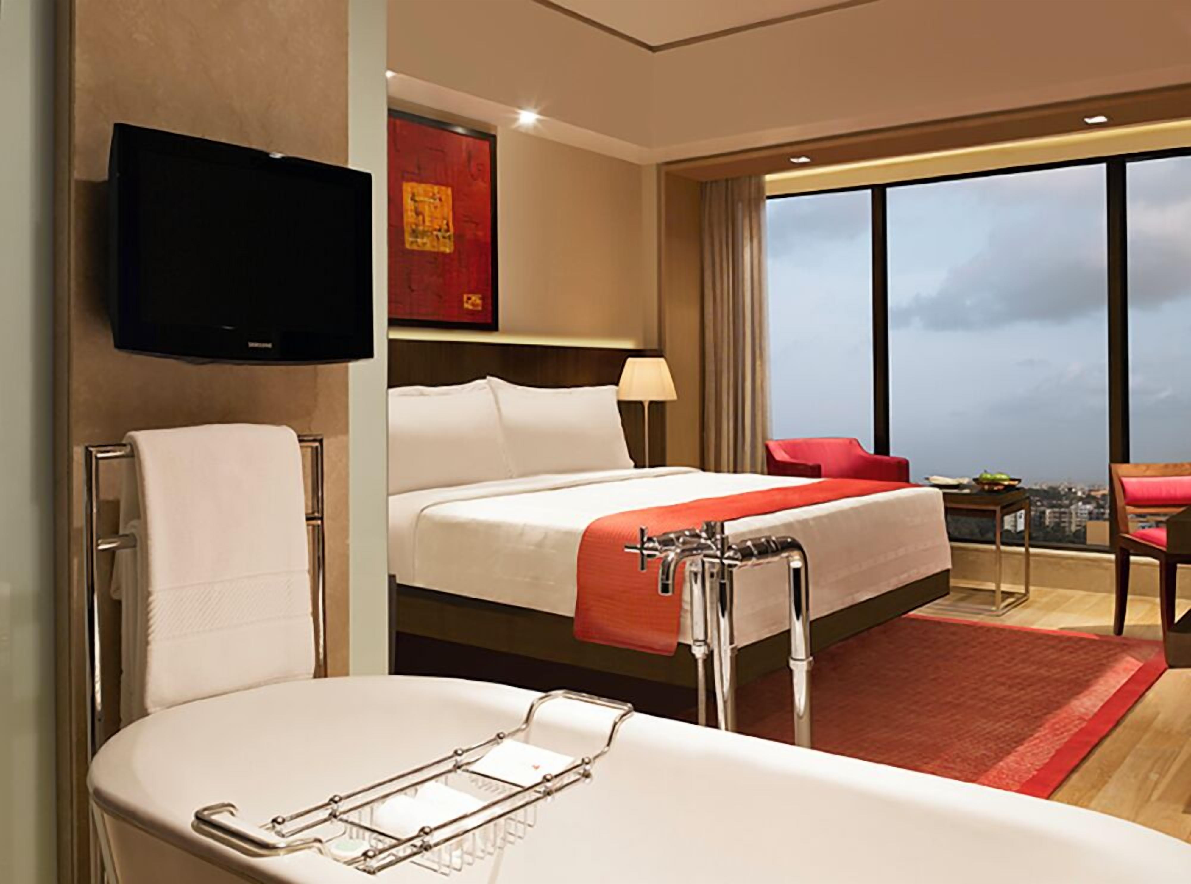 Quarto de hotel com cama grande, televisão na parede, banheira e vista para a cidade através de amplas janelas, além de uma cadeira vermelha.