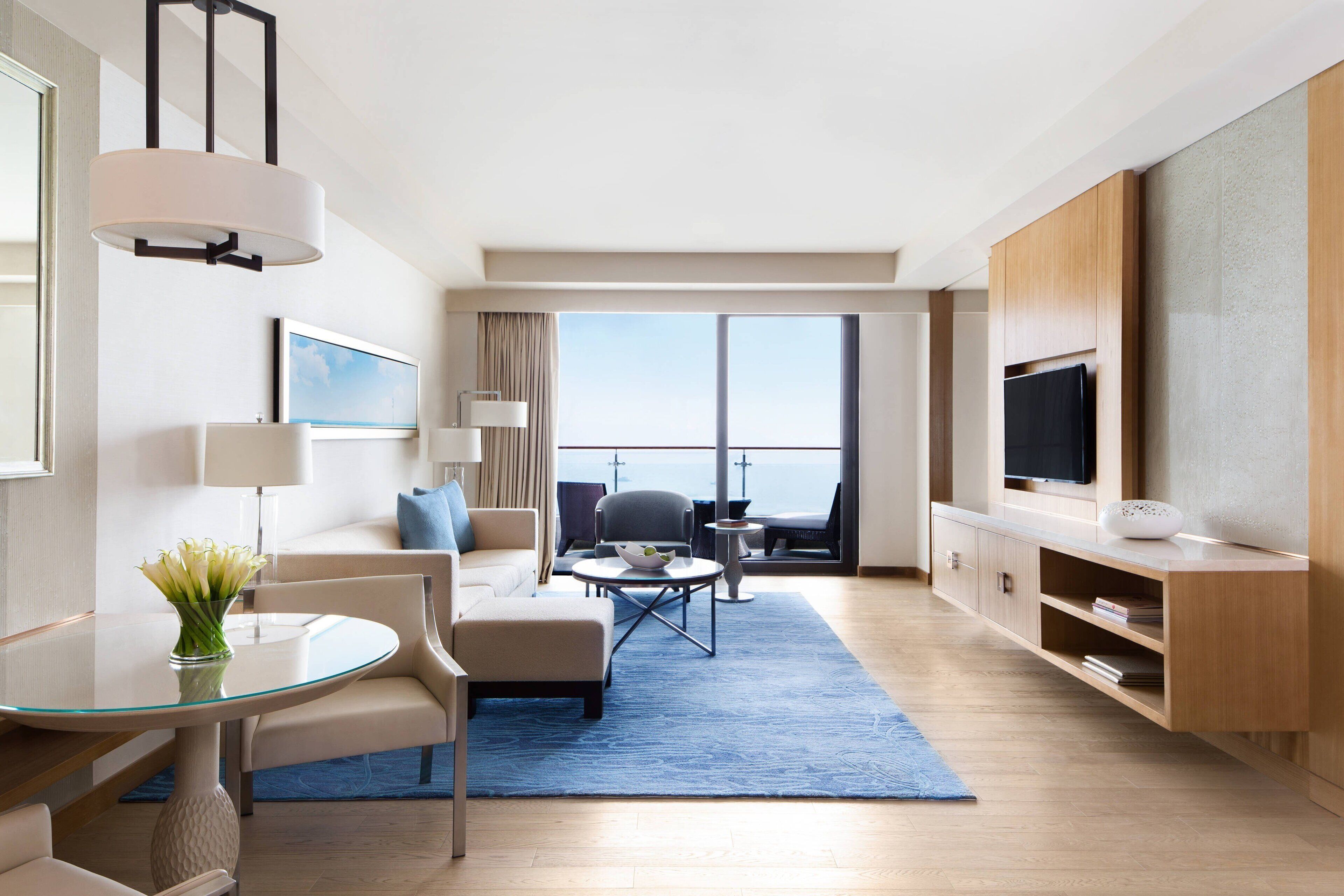 Quarto de hotel moderno com mesa de jantar de vidro, sofá, mesa de centro e televisão em um armário de madeira.
