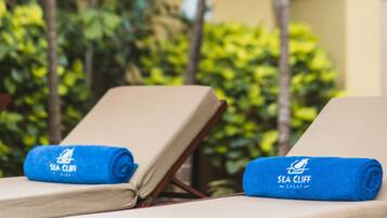 Uma área junto à piscina com espreguiçadeiras e guarda-sóis, uma toalha azul com a marca 'SEA CLIFF' e vegetação exuberante ao fundo.