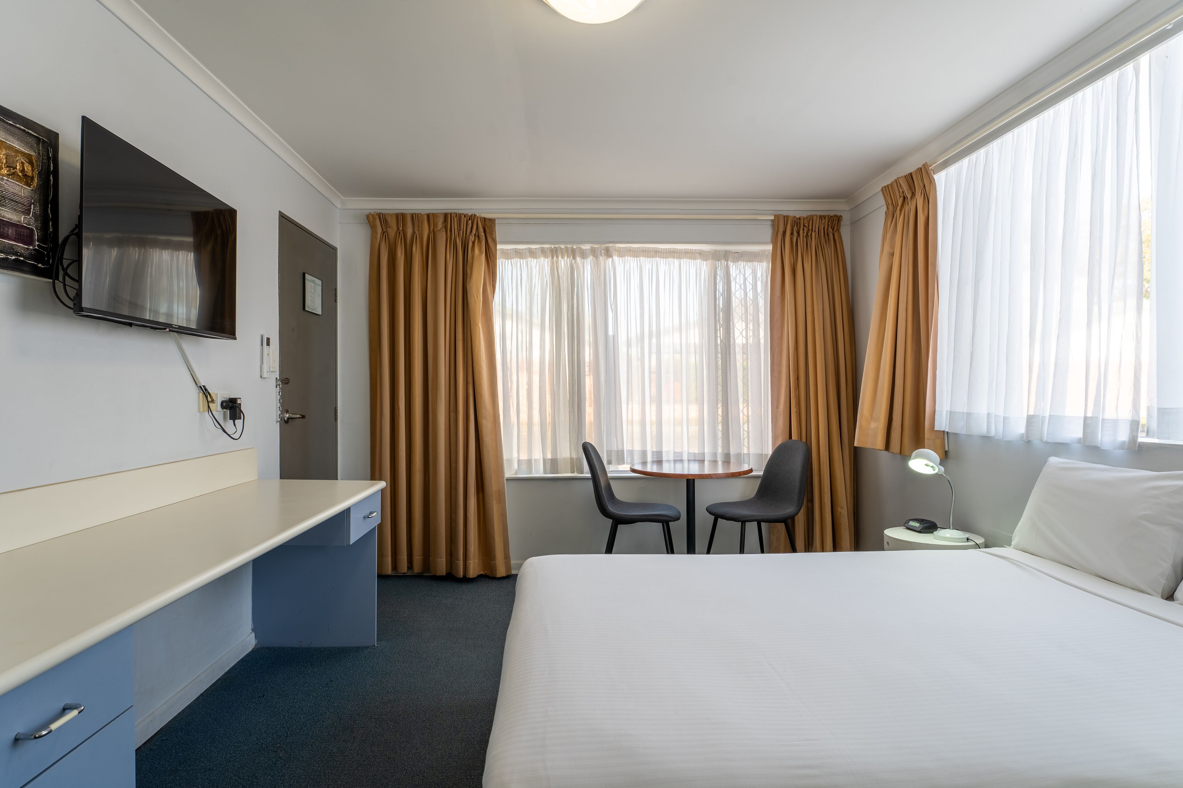 Quarto de hotel com cama, escrivaninha, duas cadeiras, televisão e janela com cortinas.