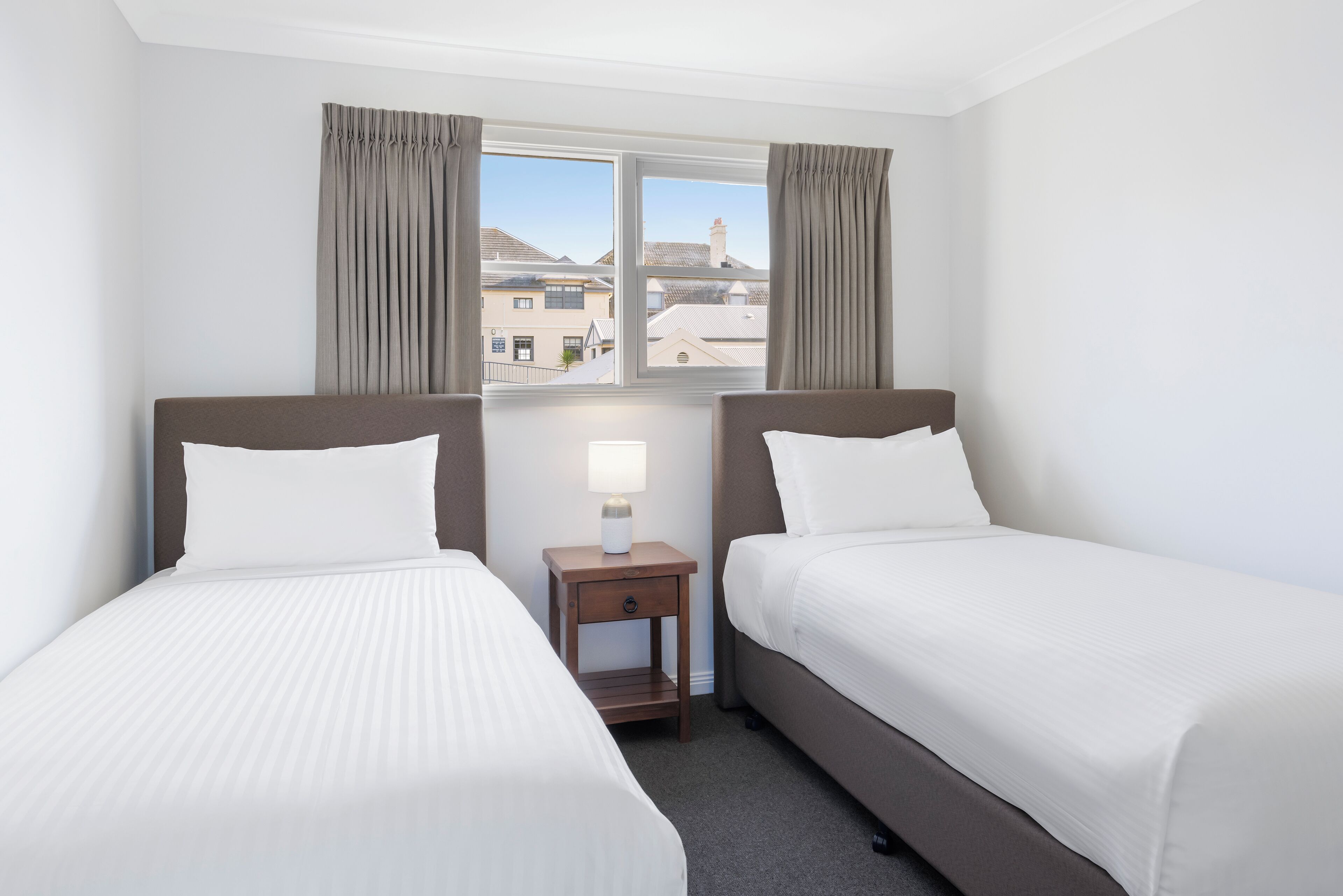 Quarto de hotel com duas camas de solteiro, uma mesa de cabeceira com abajur e uma janela com cortinas.