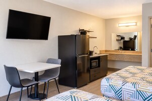 Quarto de hotel compacto com cozinha compacta, televisão de tela plana na parede, mesa de jantar com duas cadeiras e cama com roupa de cama estampada.