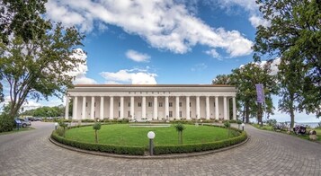 Um edifício grande, de estilo clássico, com um gramado circular central e entrada pavimentada.
