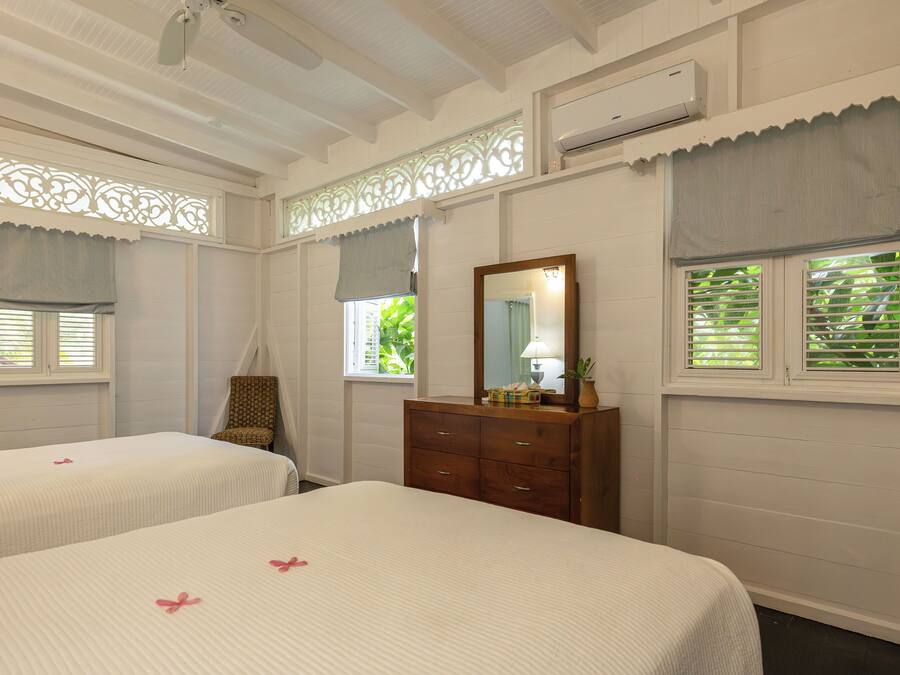 Quarto de hotel com duas camas, uma cômoda de madeira, uma cadeira, janela com persianas e ar condicionado.