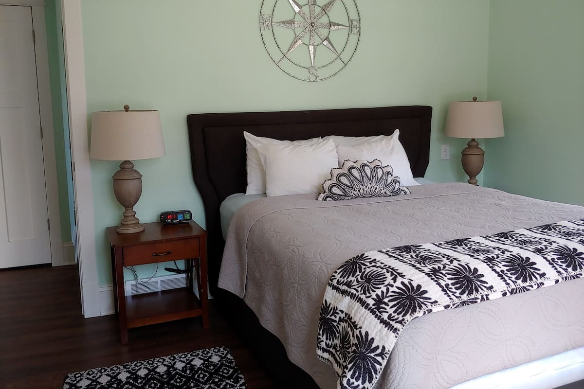 Cama bem arrumada com colcha estampada, duas luminárias de cabeceira, um objeto decorativo na parede e uma pequena mesa de cabeceira de madeira com abajur.