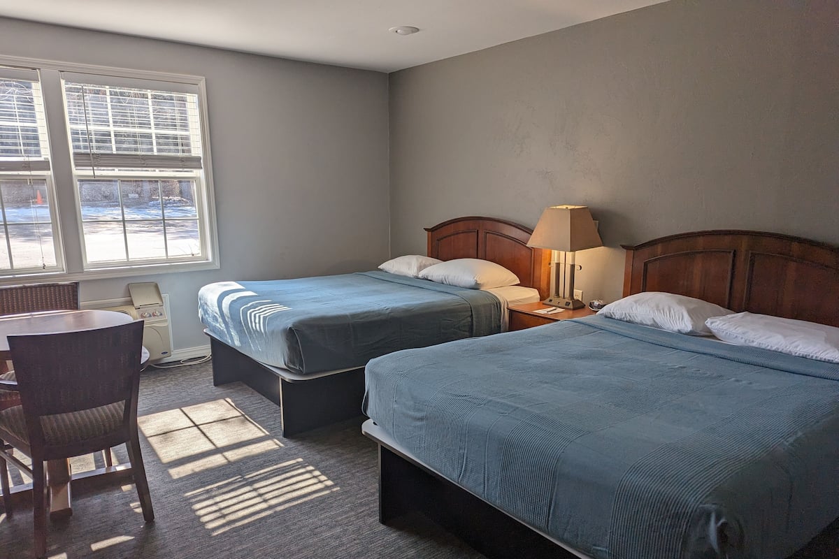 Quarto de hotel com duas camas, um painel de cabeceira em madeira, uma mesa com uma cadeira e uma janela com persianas.