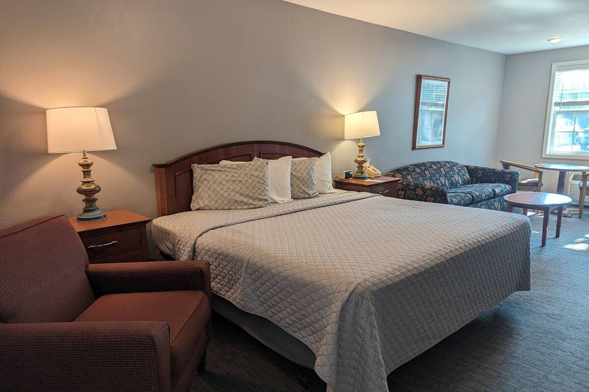 Quarto de hotel com uma cama grande, duas mesas de cabeceira com abajures, uma poltrona, um sofá e uma pequena mesa com uma cadeira.