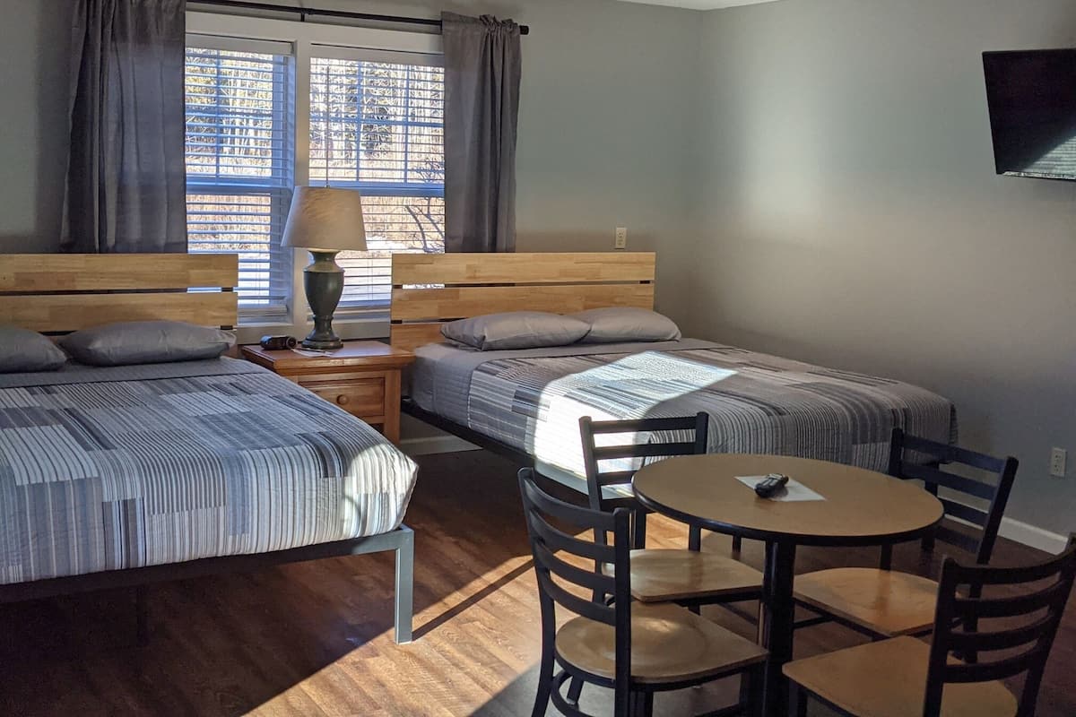 Quarto com duas camas, uma mesa e cadeiras, um abajur e uma janela com cortinas.