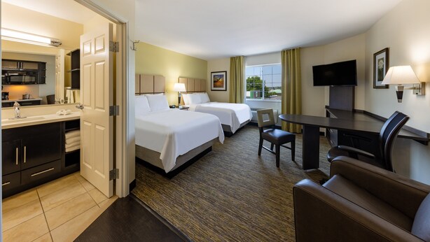 Quarto de hotel com duas camas, uma escrivaninha, uma cadeira, televisão e banheiro com pia e espelho.
