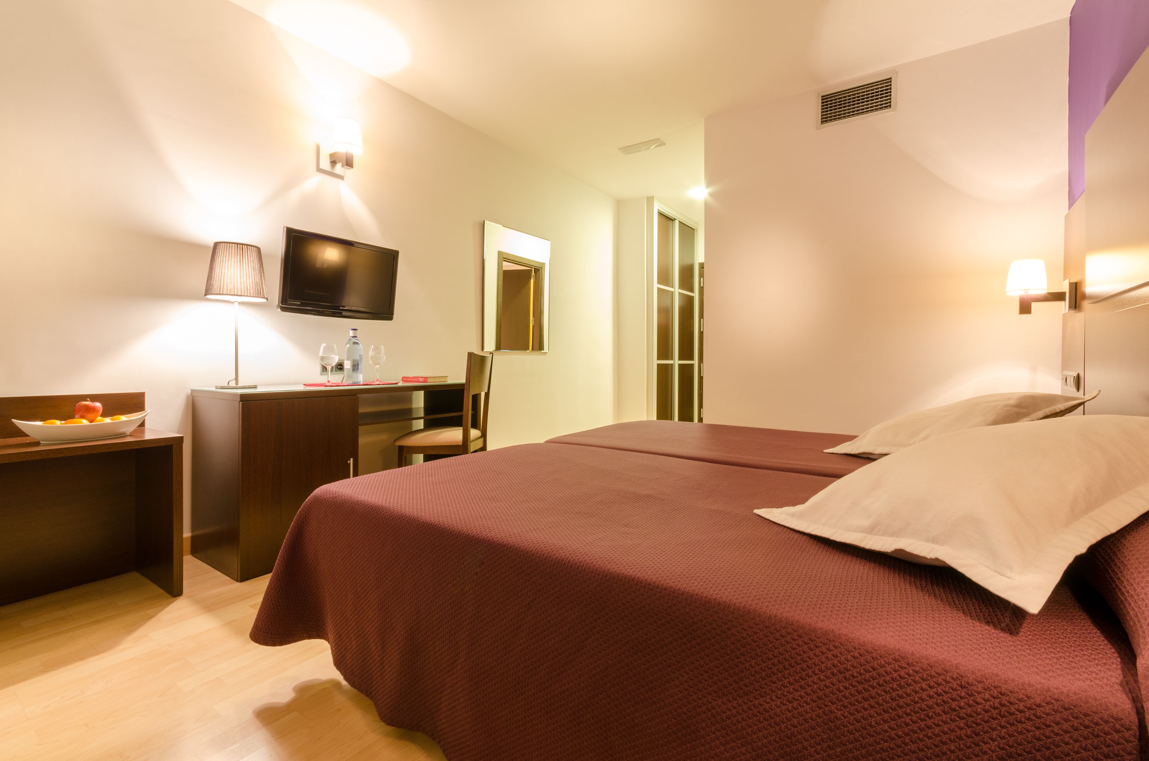 Quarto de hotel com cama, escrivaninha, cadeira, televisão e espelho.