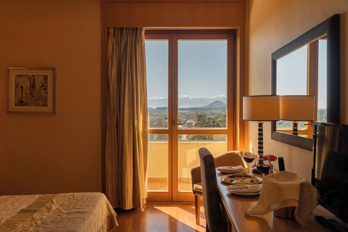 Quarto de hotel com cama, mesa de jantar com pratos e taça de vinho, uma cadeira, abajur, uma janela grande com cortinas e um quadro na parede.