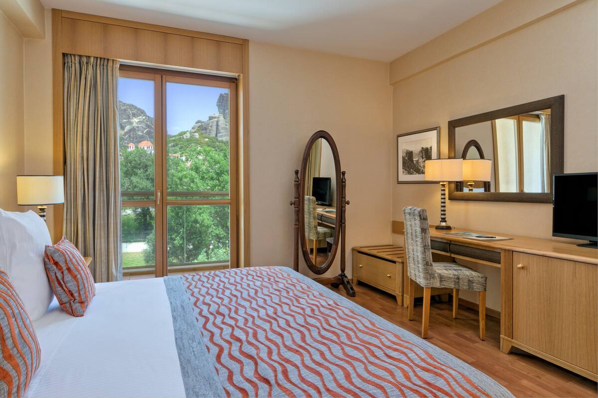 Quarto de hotel com cama, escrivaninha, cadeira, televisão, espelho e janela com vista para uma montanha.