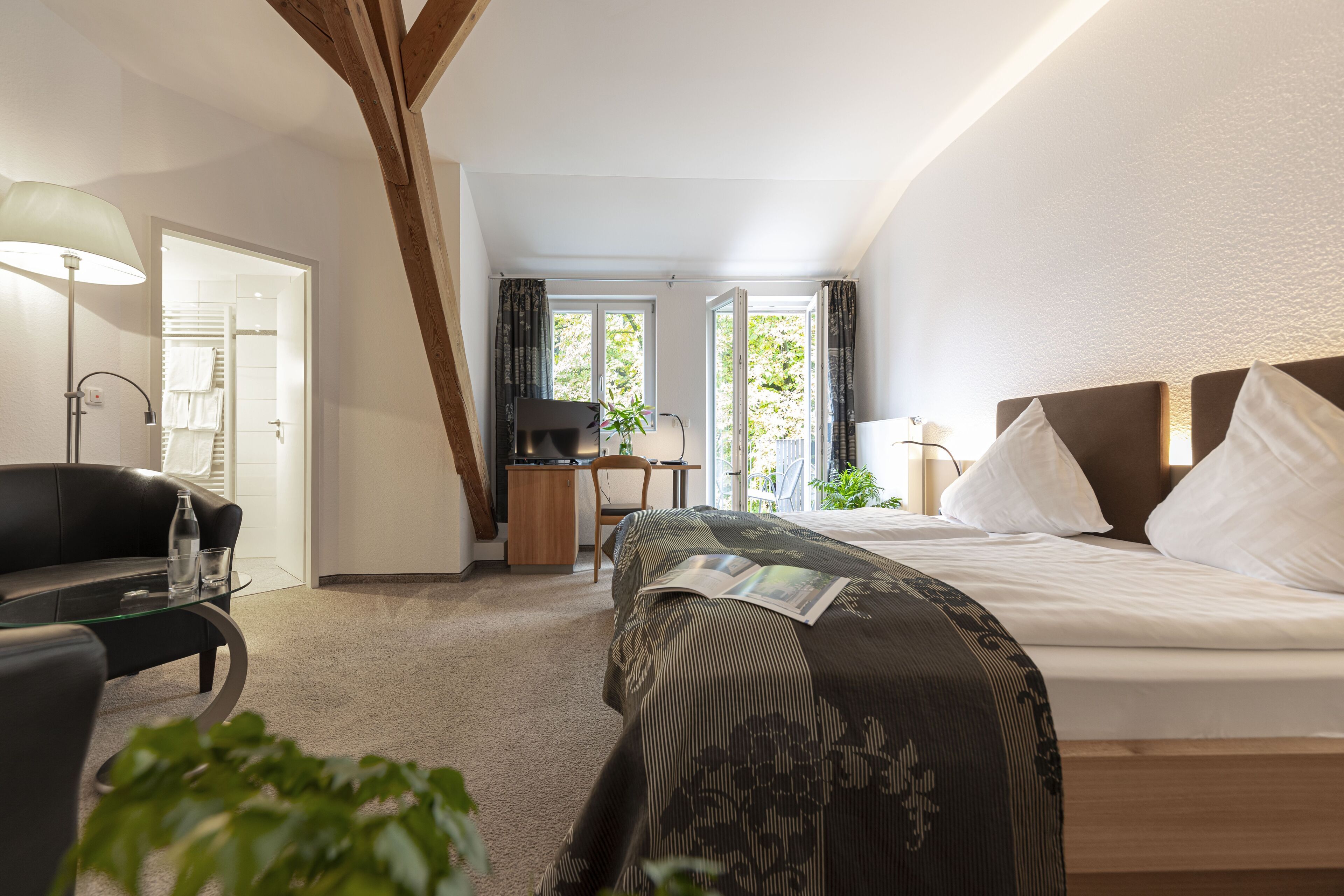 Quarto de hotel moderno com cama, escrivaninha, cadeira e sofá. Há uma janela com cortinas e uma planta na escrivaninha.