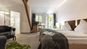 Quarto de hotel moderno com cama, escrivaninha, cadeira e sofá. Há uma janela com cortinas e uma planta na escrivaninha.