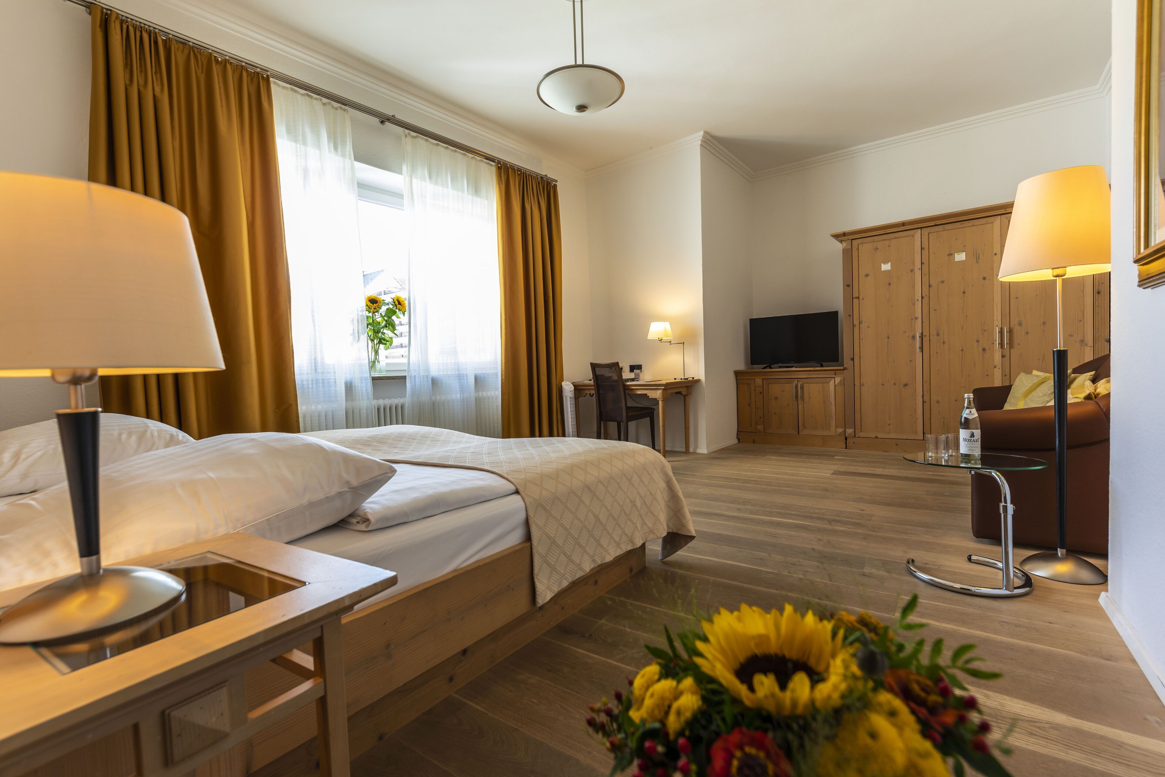 Quarto de hotel com cama, mesa de cabeceira, luminária, televisão, guarda-roupa de madeira e escrivaninha com cadeira.