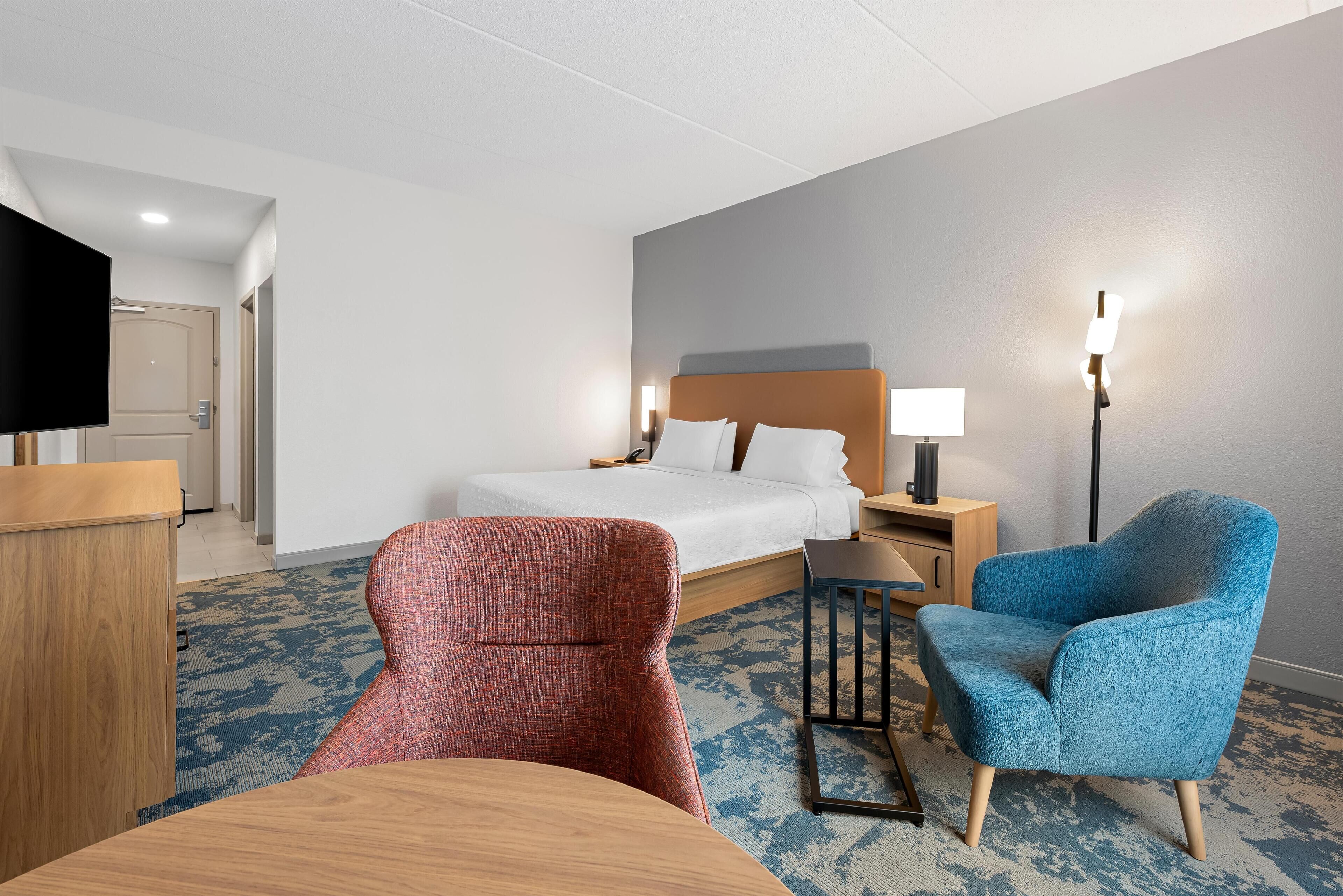 Quarto de hotel com cama, duas poltronas, uma escrivaninha e televisão.