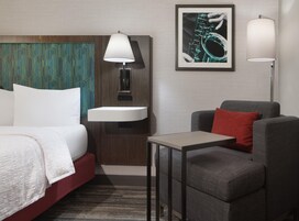 Quarto de hotel com cama, mesa de cabeceira, cadeira, mesinha e pia fixada na parede, além de um quadro emoldurado.