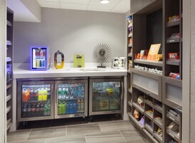 Uma estação de bebidas com serviço próprio, com dispensadores refrigerados, uma placa de neon e uma prateleira com lanches.