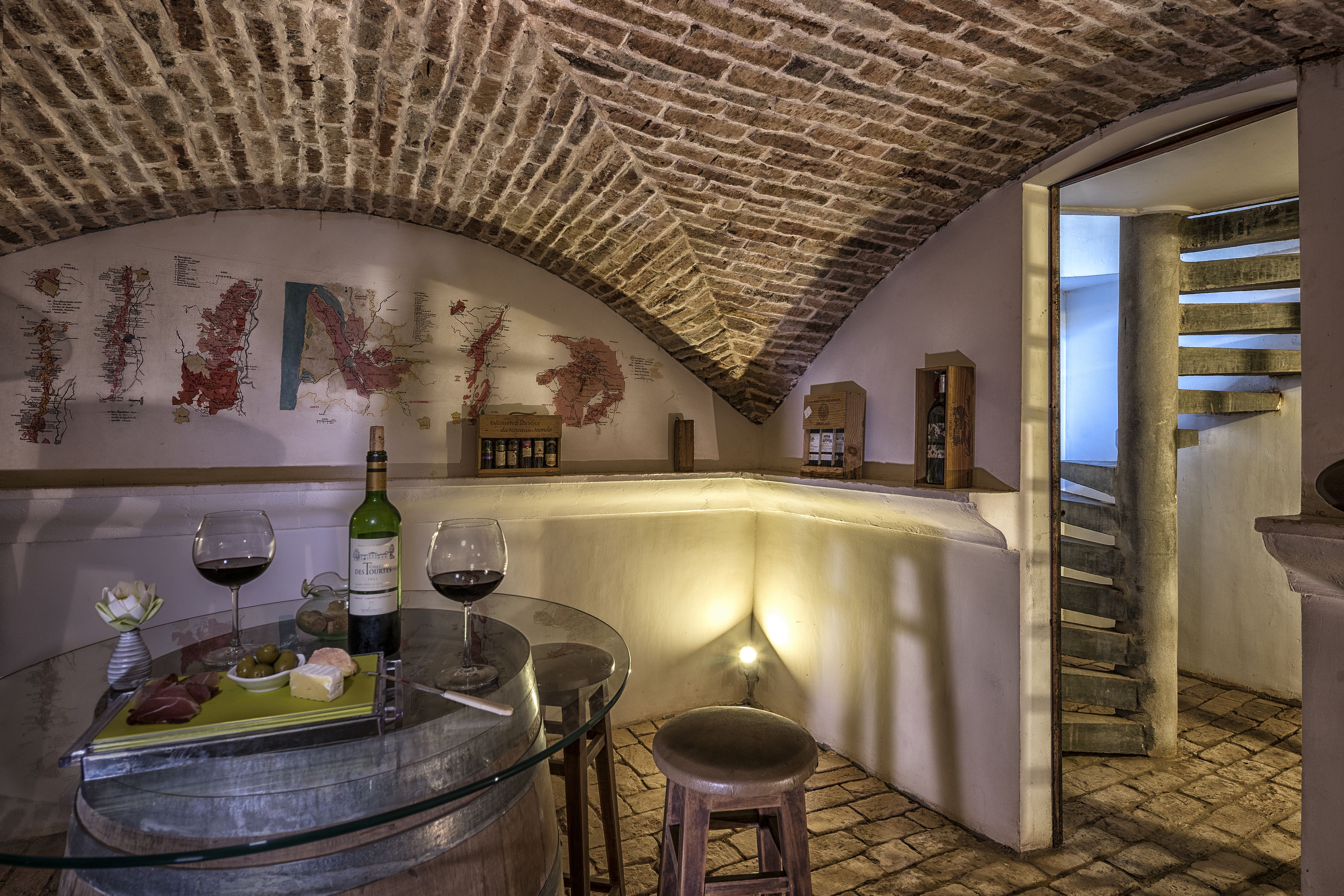 Uma área de degustação de vinhos com uma mesa de vidro, garrafas e copos de vinho. Possui teto de tijolos e uma escada ao fundo.
