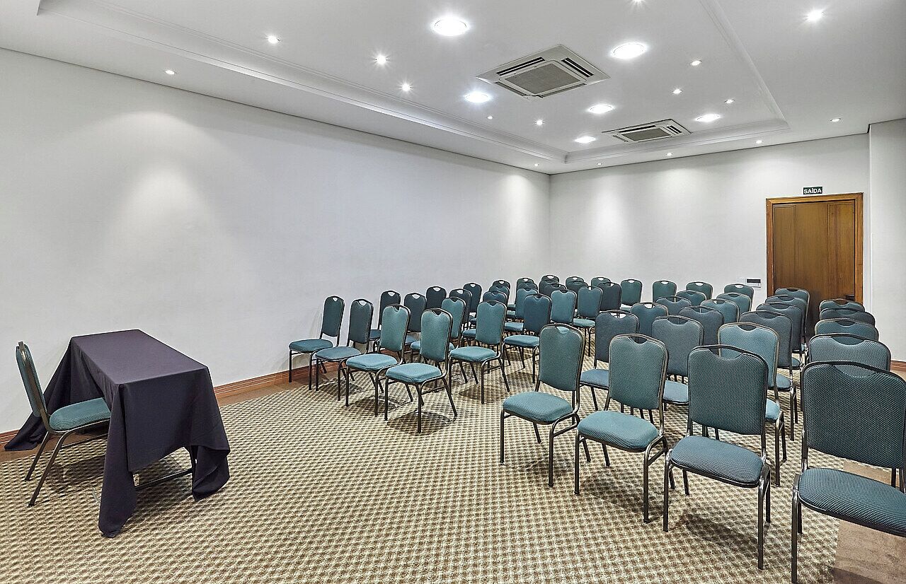 Uma sala de conferências com fileiras de cadeiras, uma mesa e uma porta com a inscrição 'BAIDA'.