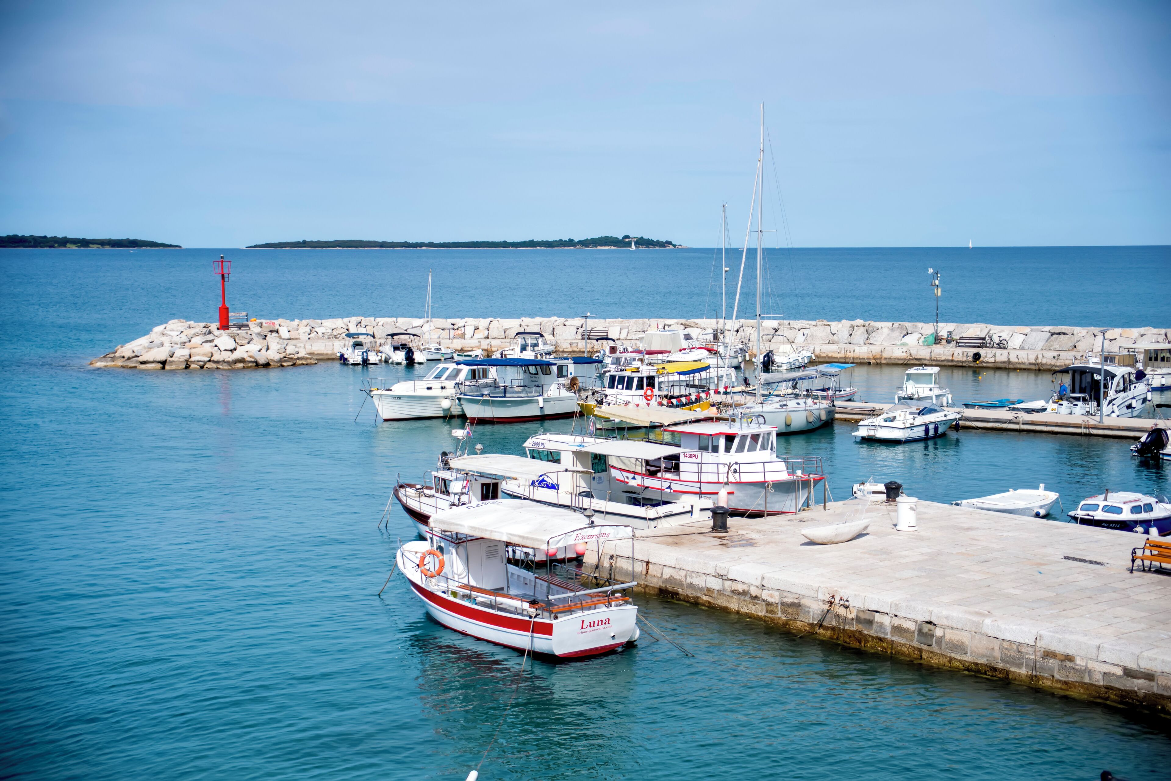 Marina com diversos barcos atracados, um farol ao longe e céu claro.