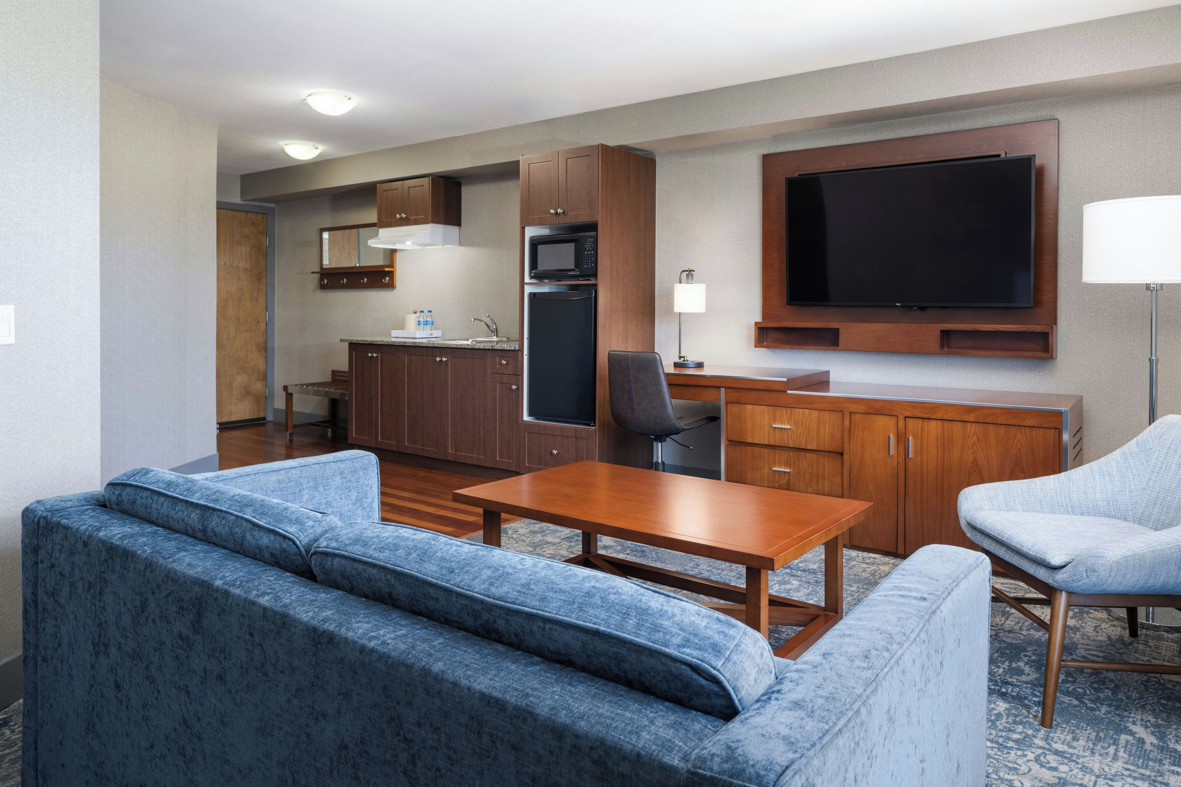 Quarto de hotel com cozinha compacta, televisão de tela plana na parede, sofá, mesa de jantar e luminária.