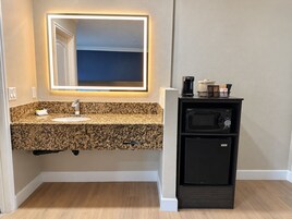 Casa de banho com bancada de granito, lavatório, espelho iluminado por baixo e máquina de café, além de frigorífico pequeno.