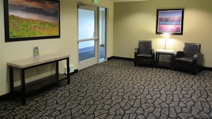 Um hall de hotel com piso coberto por carpete, duas cadeiras, uma mesa pequena e duas imagens emolduradas nas paredes.