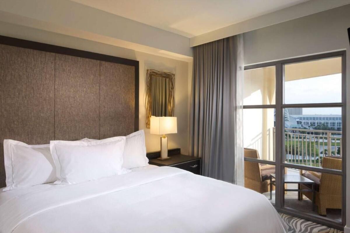 Quarto de hotel com uma cama grande, uma mesa de cabeceira com abajur, vista de um edifício pela janela e uma varanda com cadeiras.