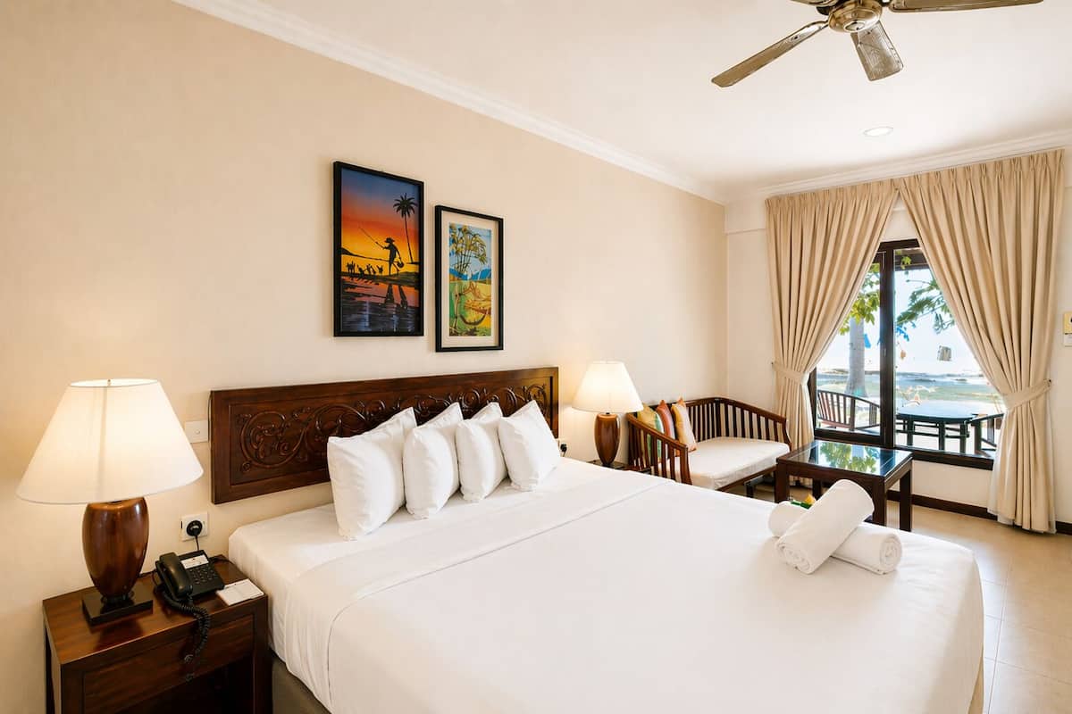 Quarto de hotel com uma cama grande, duas luminárias laterais, cabeceira de madeira, escrivaninha com telefone, cadeira e vista para o mar pela janela.