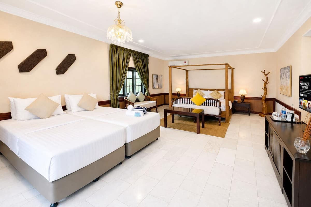 Quarto de hotel com duas camas, um espaço para sentar, um banco de madeira, um candelabro e uma televisão fixada na parede.