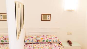 Quarto com cama coberta por um edredom colorido e estampado, mesa de cabeceira com abajur e luminária na parede.