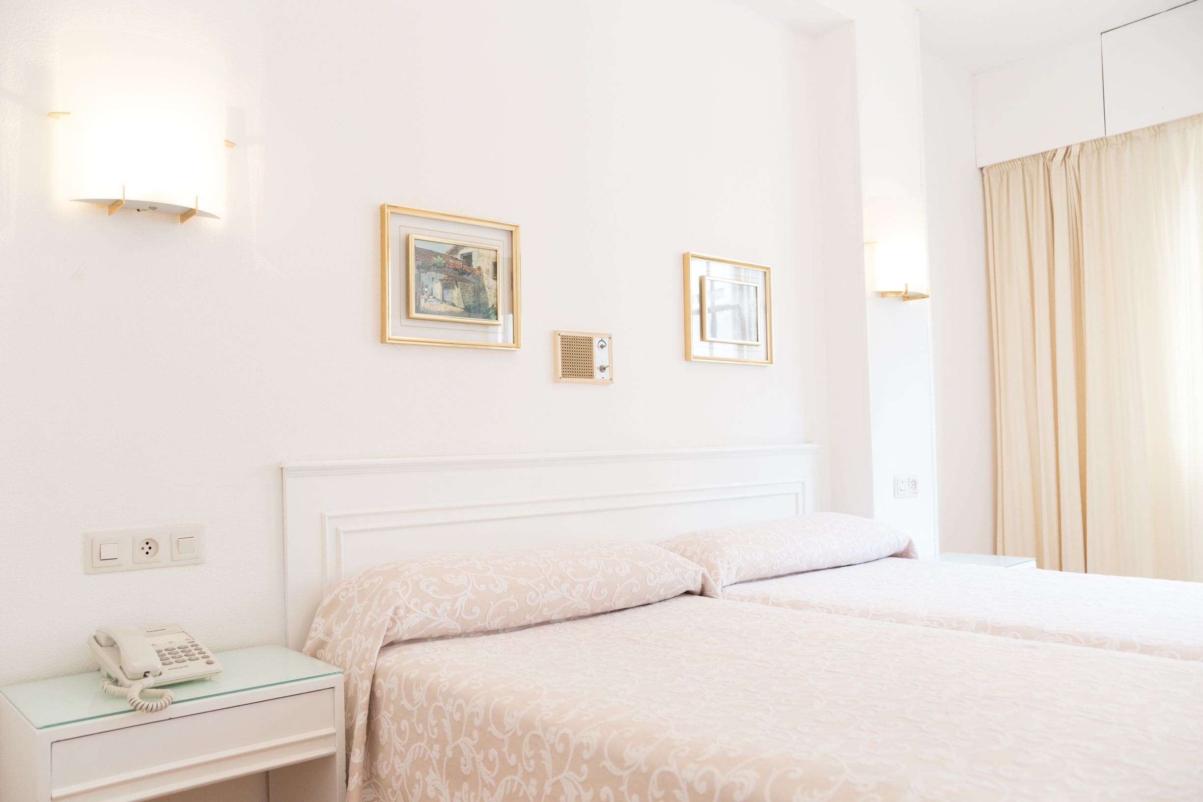 Quarto de hotel com cama, mesa de cabeceira com telefone e duas imagens emolduradas na parede.