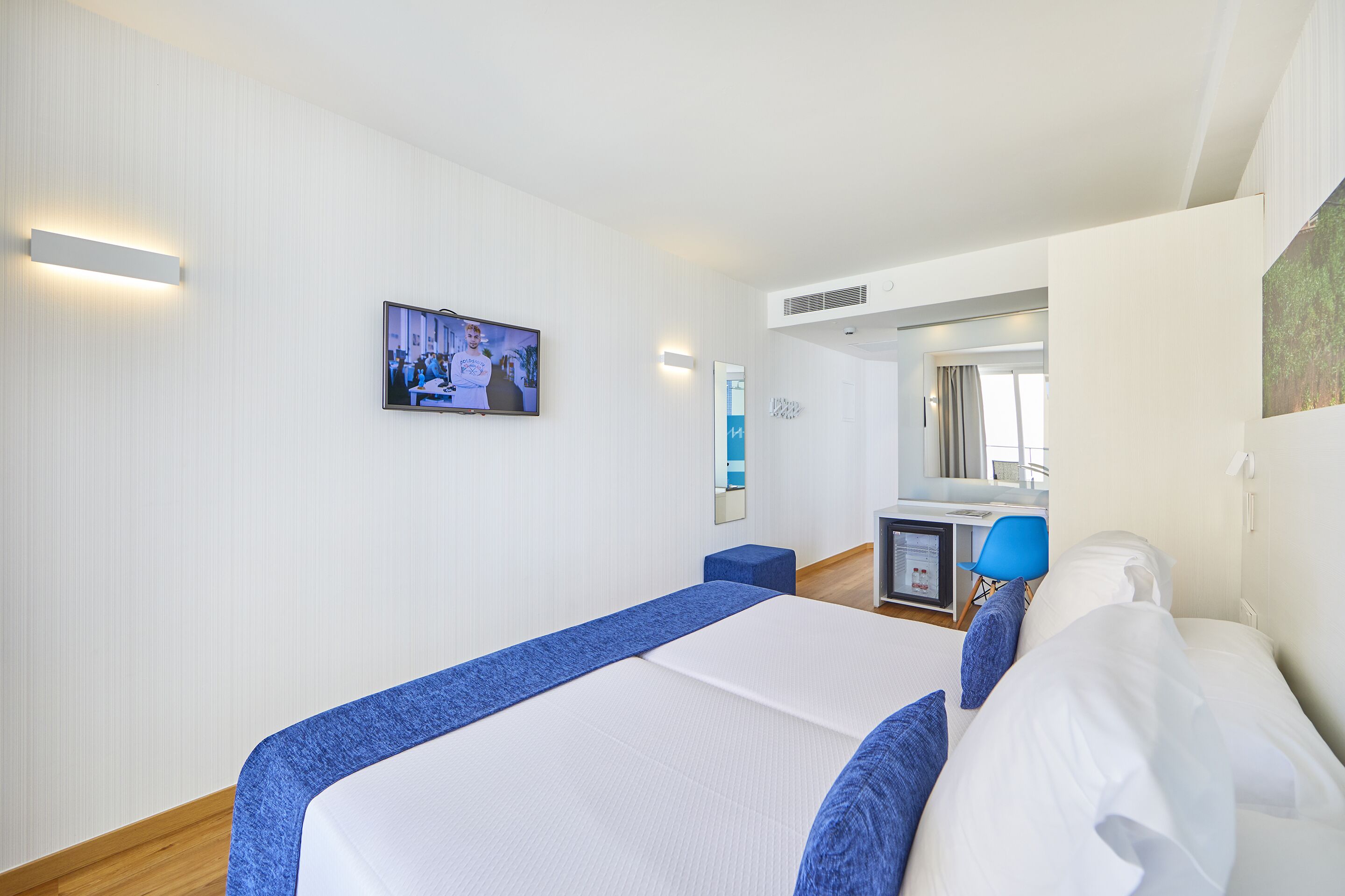 Quarto de hotel com uma cama grande, uma mesa com computador, uma televisão na parede e uma janela com cortinas.
