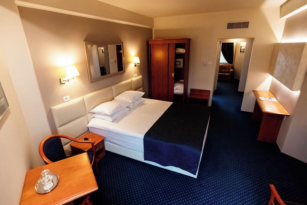 Quarto de hotel com cama, mesas de cabeceira, escrivaninha, cadeira, guarda-roupa, espelho e corredor.