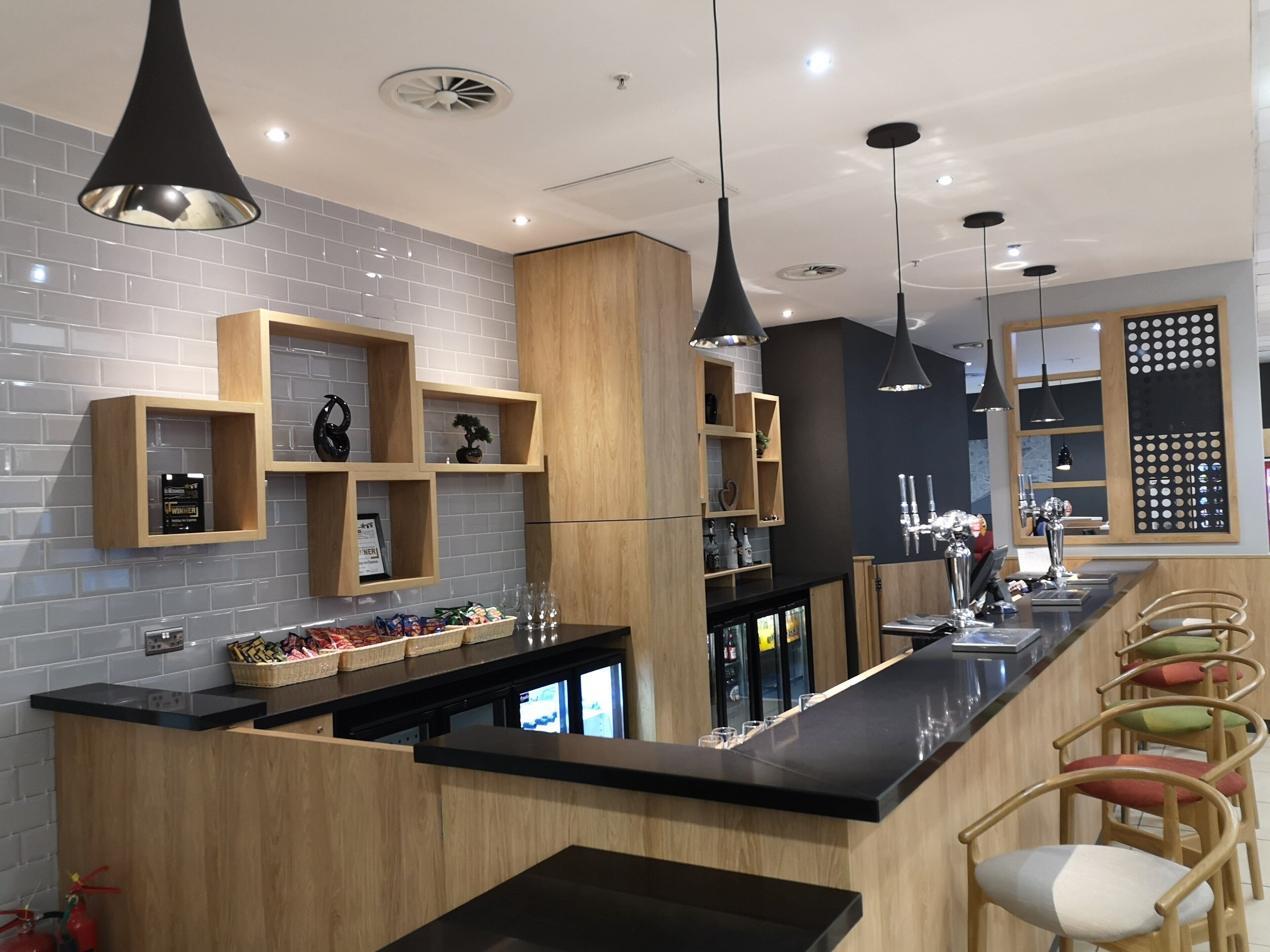 Uma área de bar moderna com prateleiras de madeira, bancada preta e iluminação pendente.