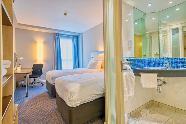 Quarto de hotel com duas camas, uma mesa, uma cadeira, banheiro com pia e espelho, e vista para o exterior.