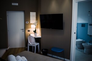 Quarto de hotel com uma cama, escrivaninha com cadeira, televisão e banheiro visível através de uma porta aberta.