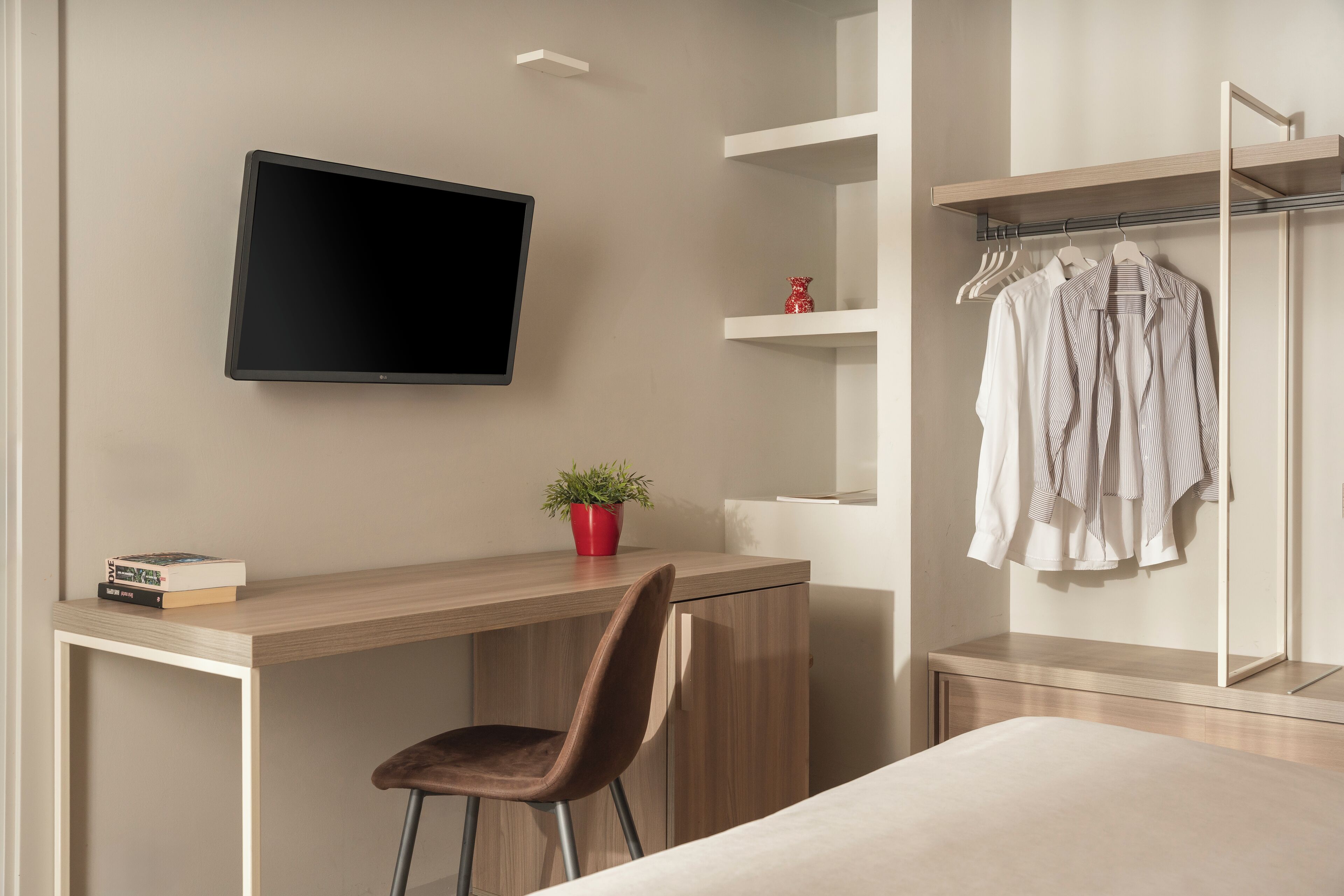 Quarto moderno com escrivaninha de madeira, uma cadeira, uma cama, televisão, prateleira com roupas e uma pequena planta.