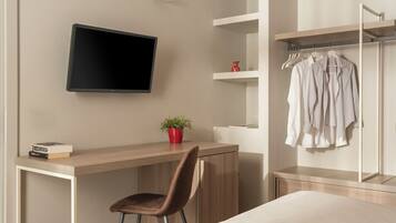 Quarto moderno com escrivaninha de madeira, uma cadeira, uma cama, televisão, prateleira com roupas e uma pequena planta.