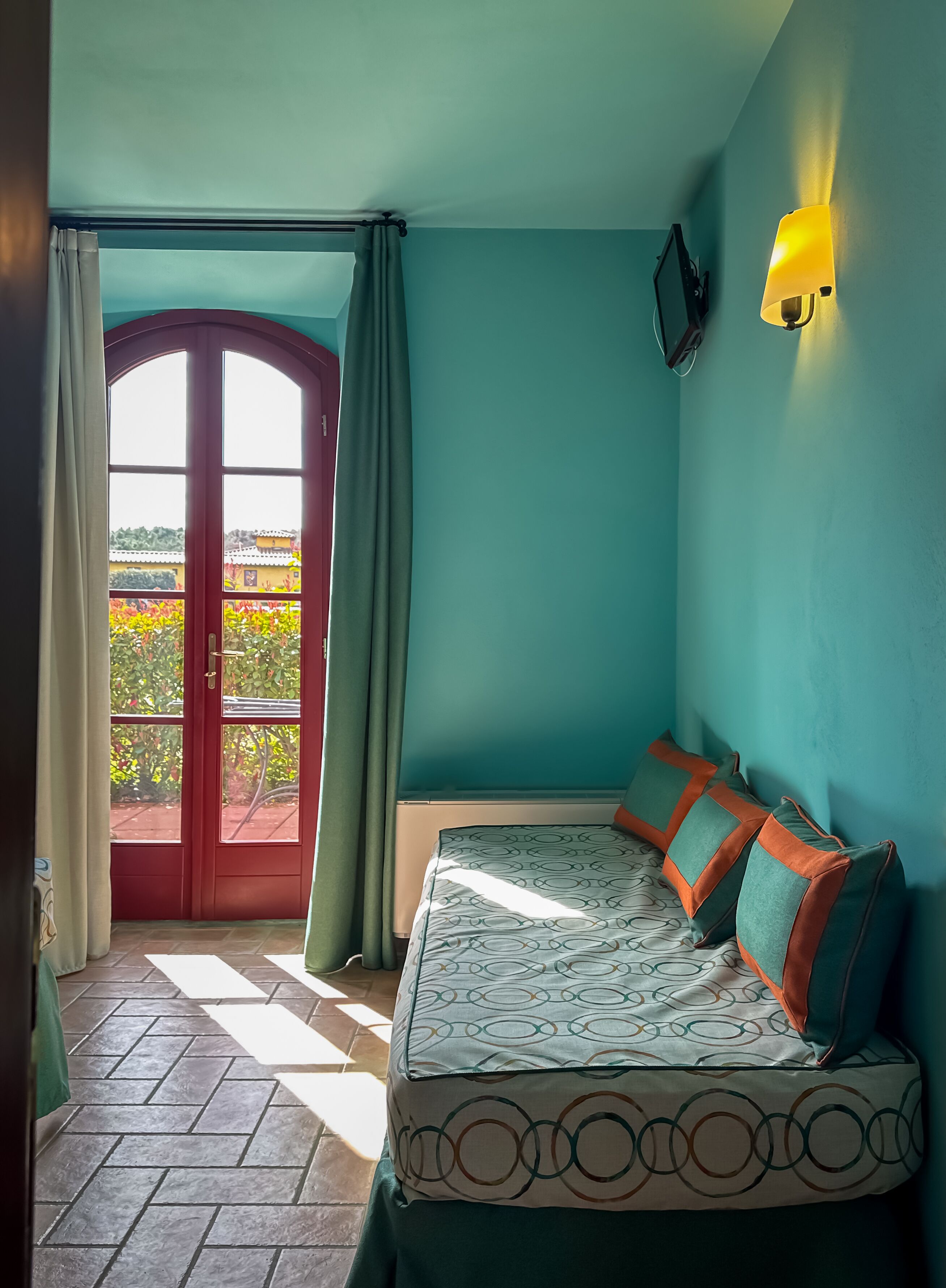 Quarto com cama, janela com cortinas, luminária fixada na parede e vista para o verde lá fora.