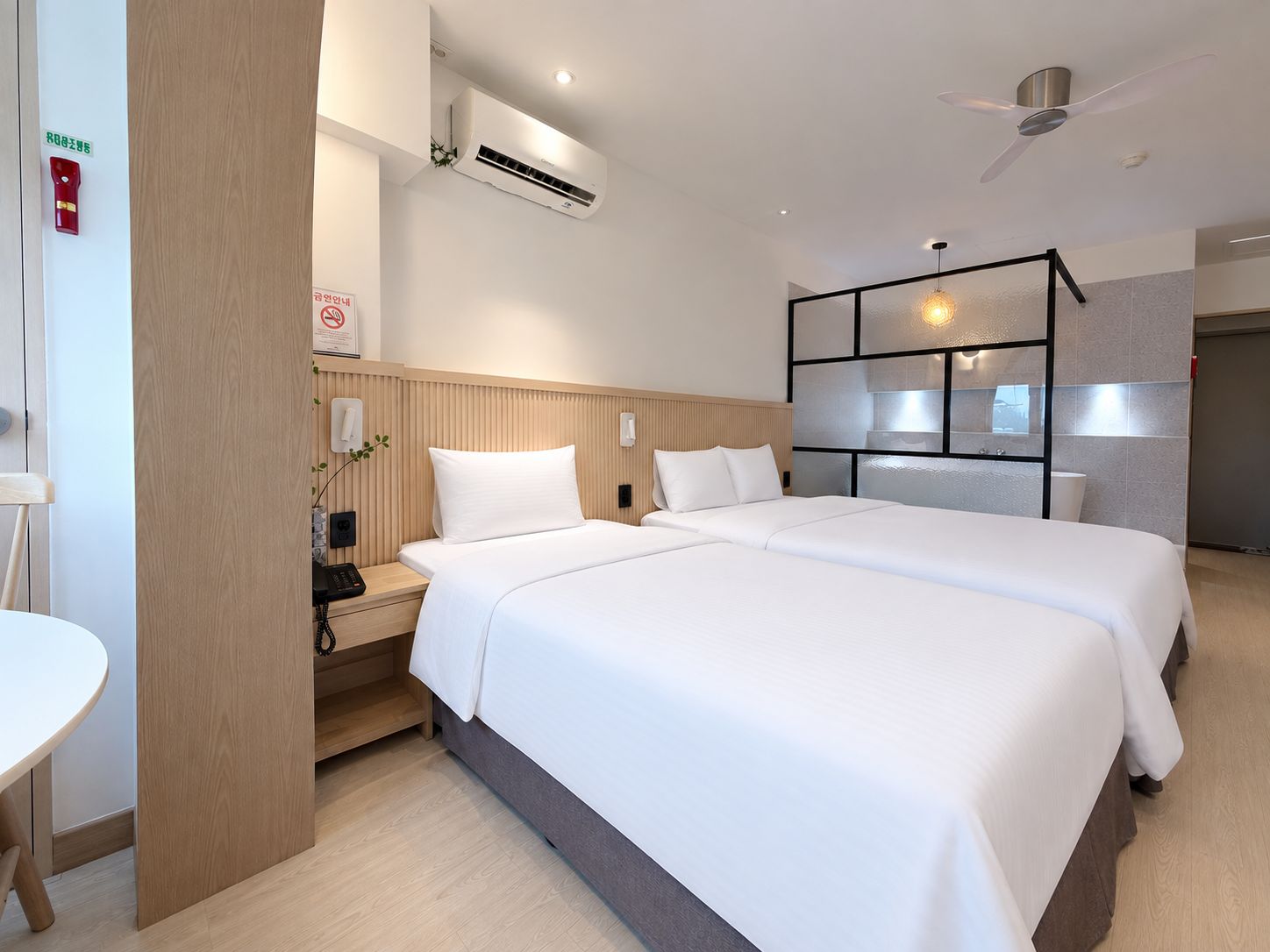 Quarto de hotel com duas camas, cabeceira de madeira, ar condicionado, escrivaninha com cadeira e uma pequena mesa.