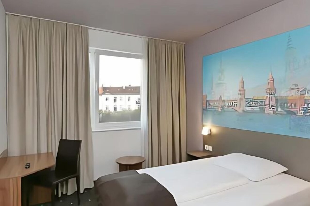 Quarto de hotel com uma cama, escrivaninha, cadeira, mesa pequena e janela com cortinas.