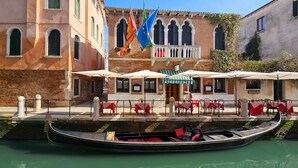 Um restaurante à beira do canal com mesas ao ar livre, uma gôndola e edifícios históricos ao fundo.