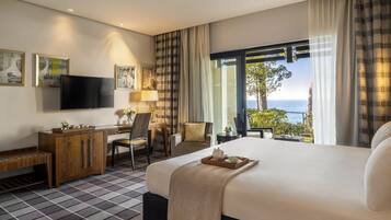 Quarto de hotel com uma cama grande, televisão, escrivaninha com cadeira e vista para o mar.