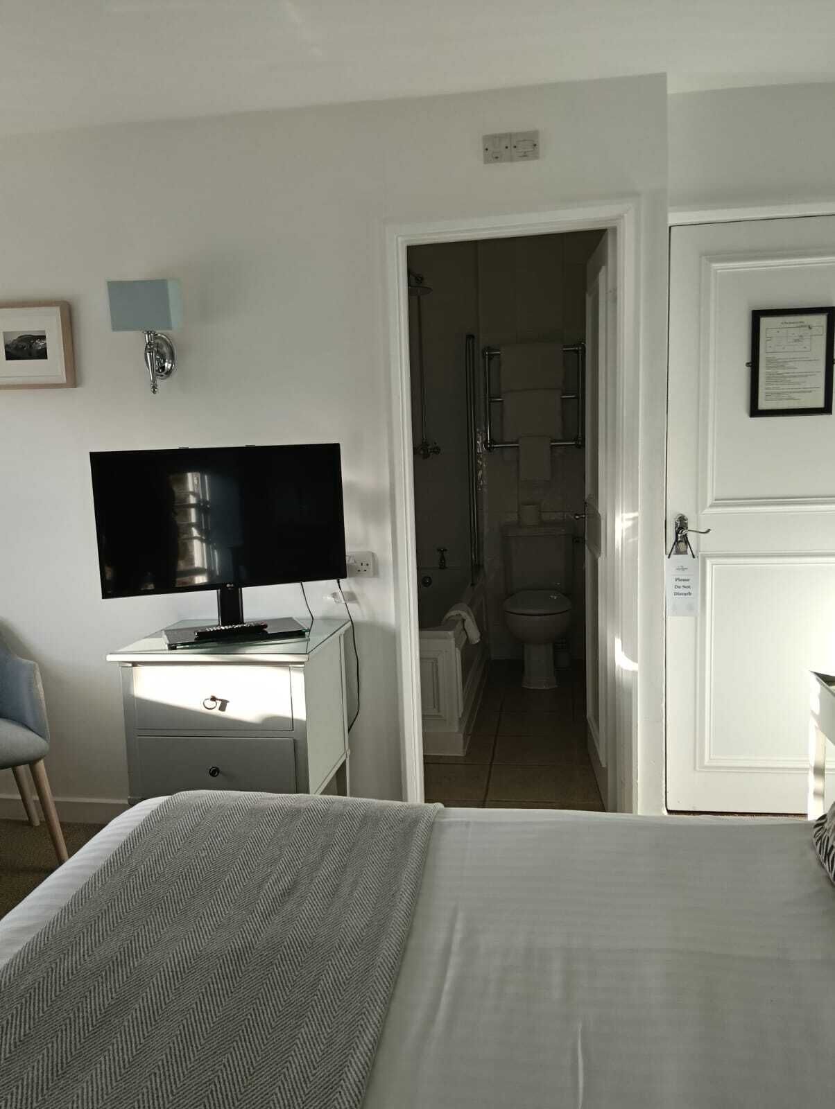 Quarto de hotel com cama, escrivaninha com televisão, área de chuveiro e porta branca.