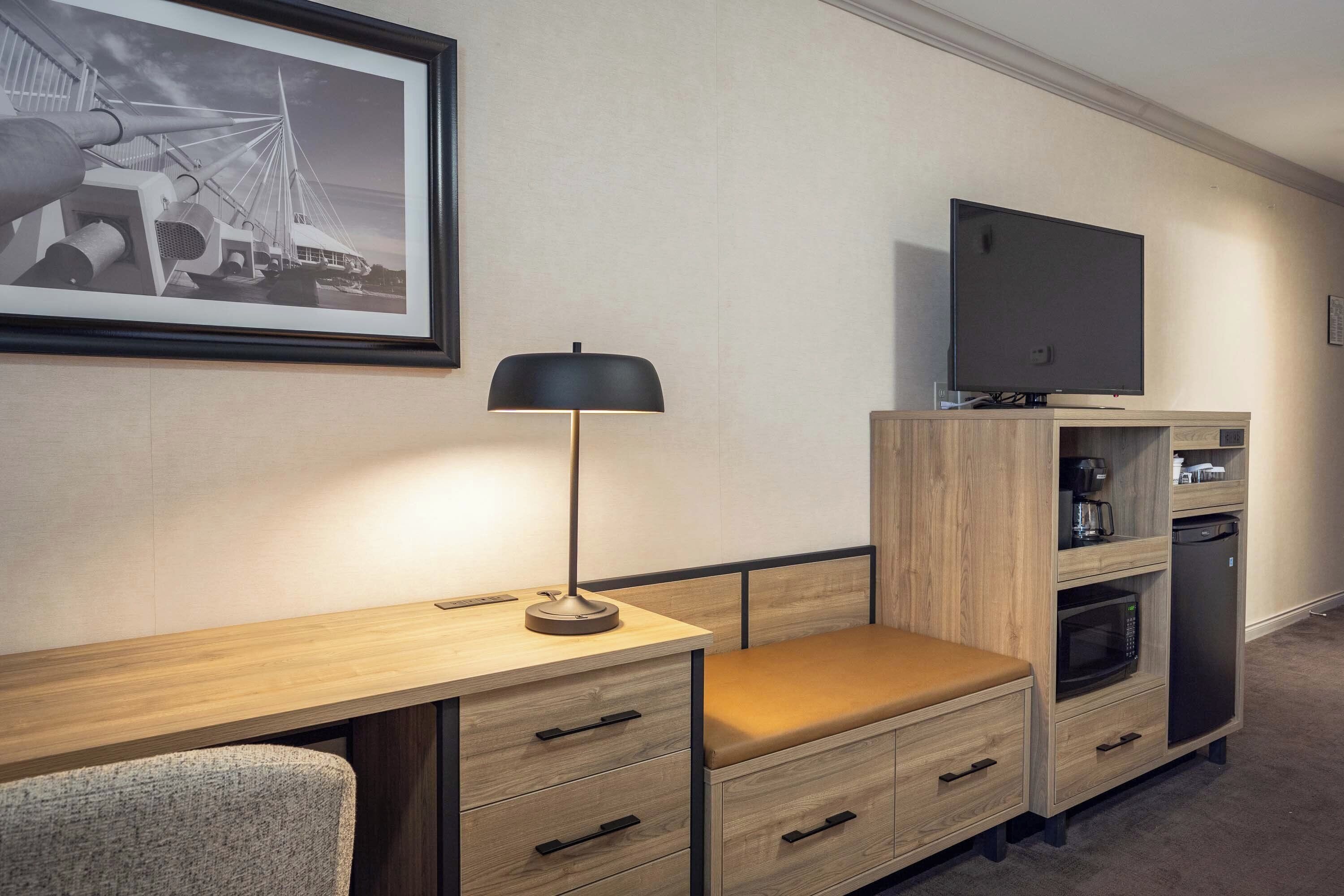 Quarto de hotel com uma escrivaninha de madeira, uma luminária preta, uma televisão sobre um suporte de madeira, um micro-ondas e uma geladeira.